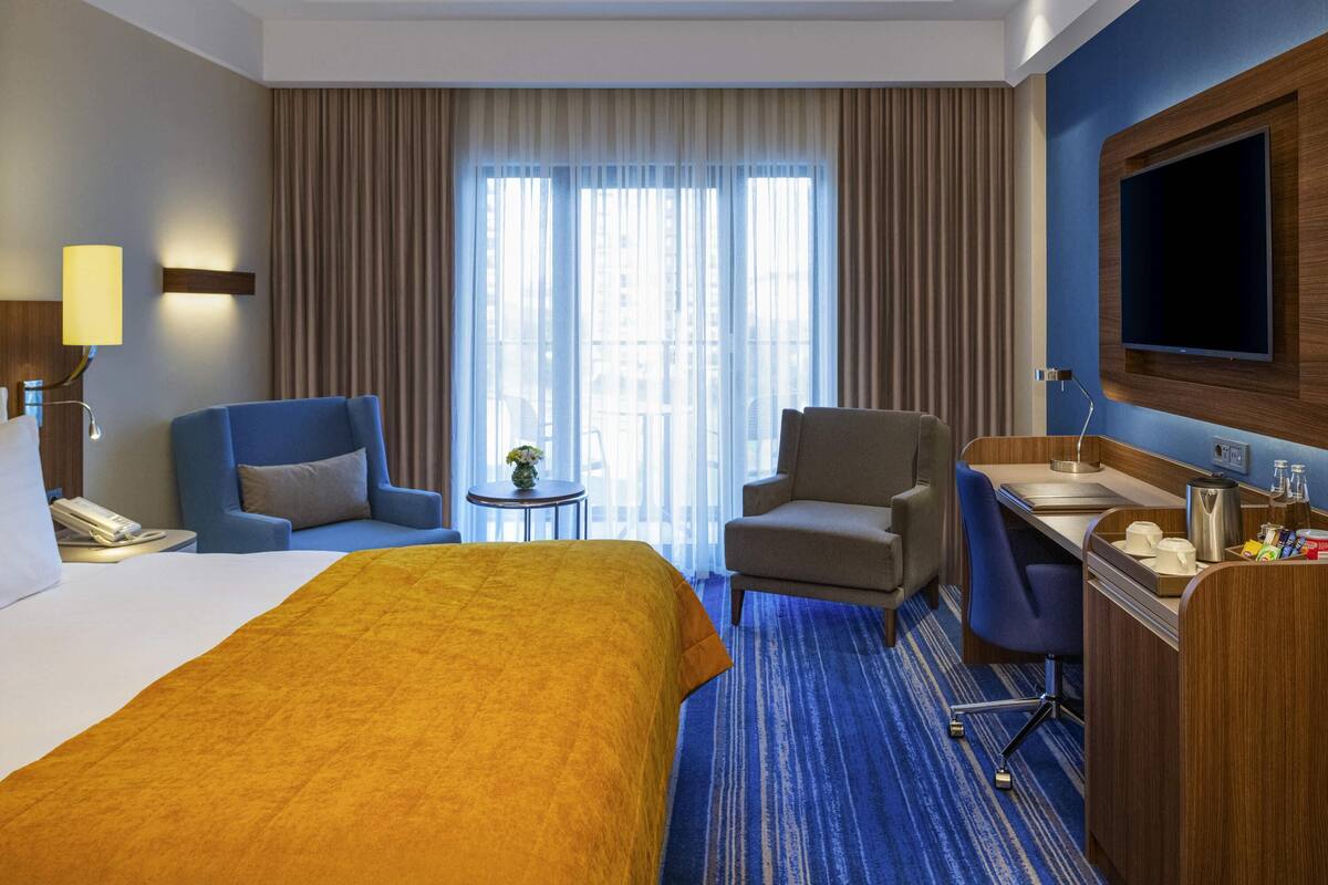 Quarto de hotel com uma cama, duas poltronas, uma escrivaninha, televisão e vista da cidade através de amplas janelas.