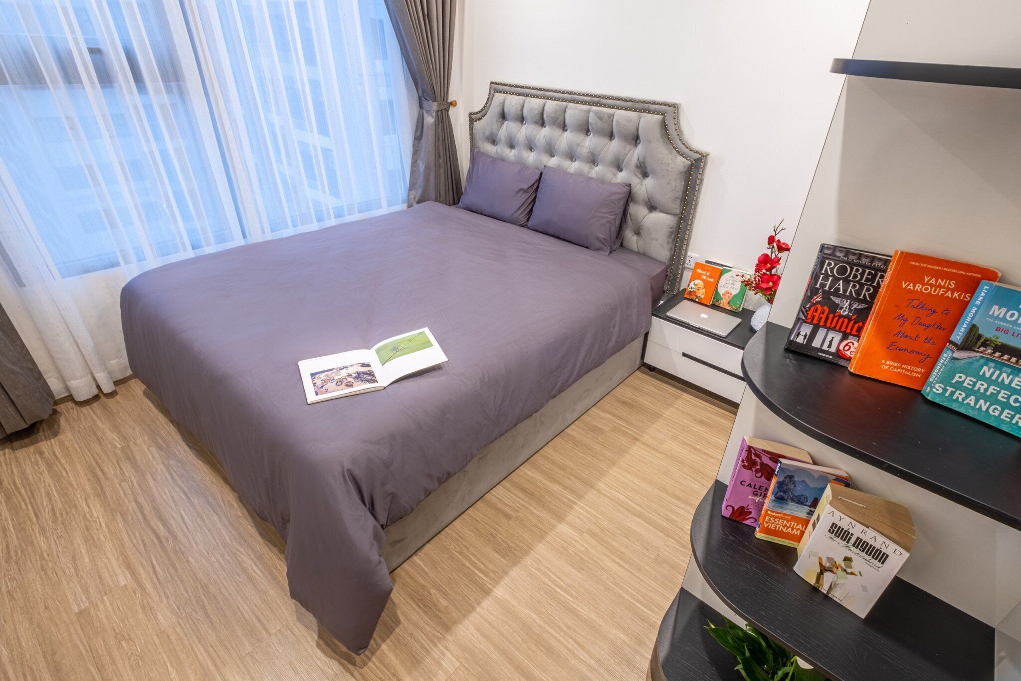 Uma cama bem arrumada com um livro em cima, uma mesinha de cabeceira com livros e um criado-mudo com uma luminária.