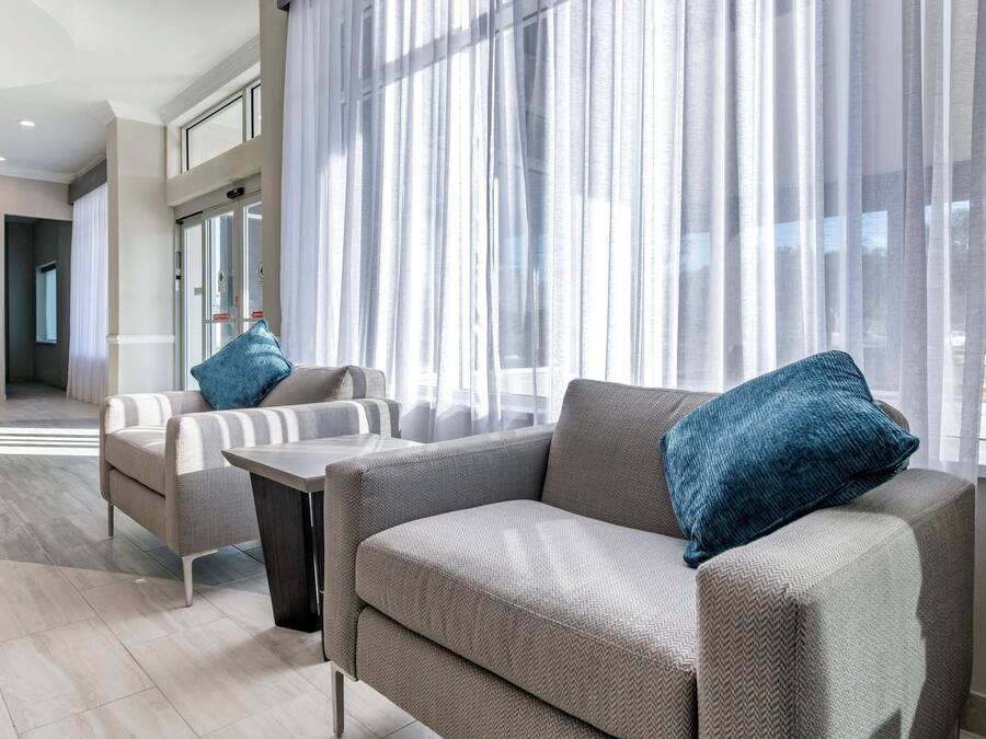 Quarto de hotel moderno com sofá e poltrona, janelas amplas com cortinas leves e piso ladrilhado.