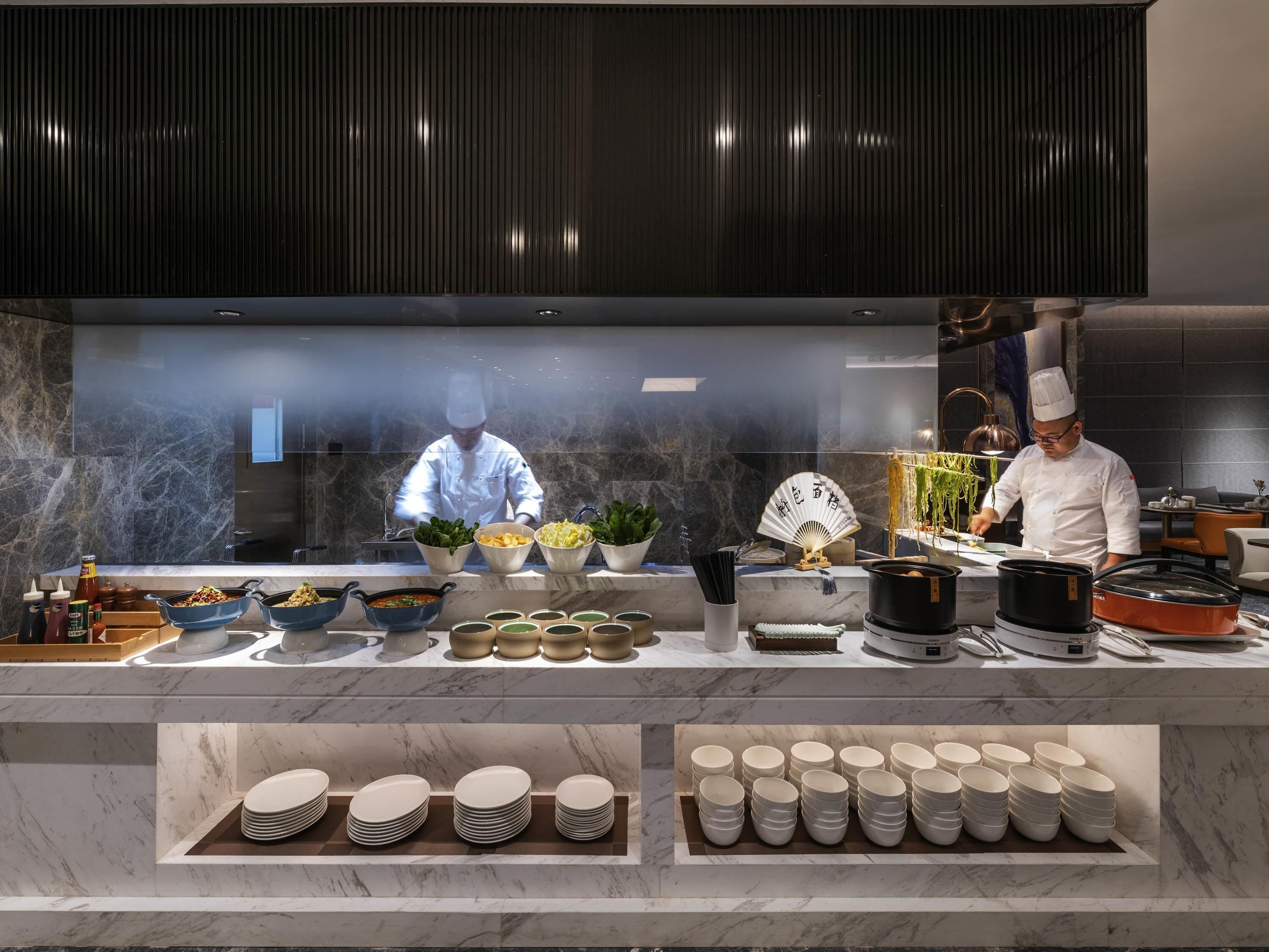 Uma cozinha moderna com chefs preparando alimentos, bancadas de mármore e diversos utensílios e ingredientes de cozinha.