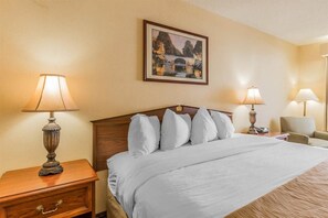 Quarto de hotel com uma cama, duas luminárias de cabeceira, um criado-mudo, uma cadeira e um quadro na parede.