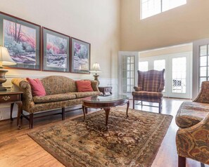 Uma sala de estar com sofá, poltrona, mesa de centro e tapete. Há quadros nas paredes, um abajur e uma janela com vista.