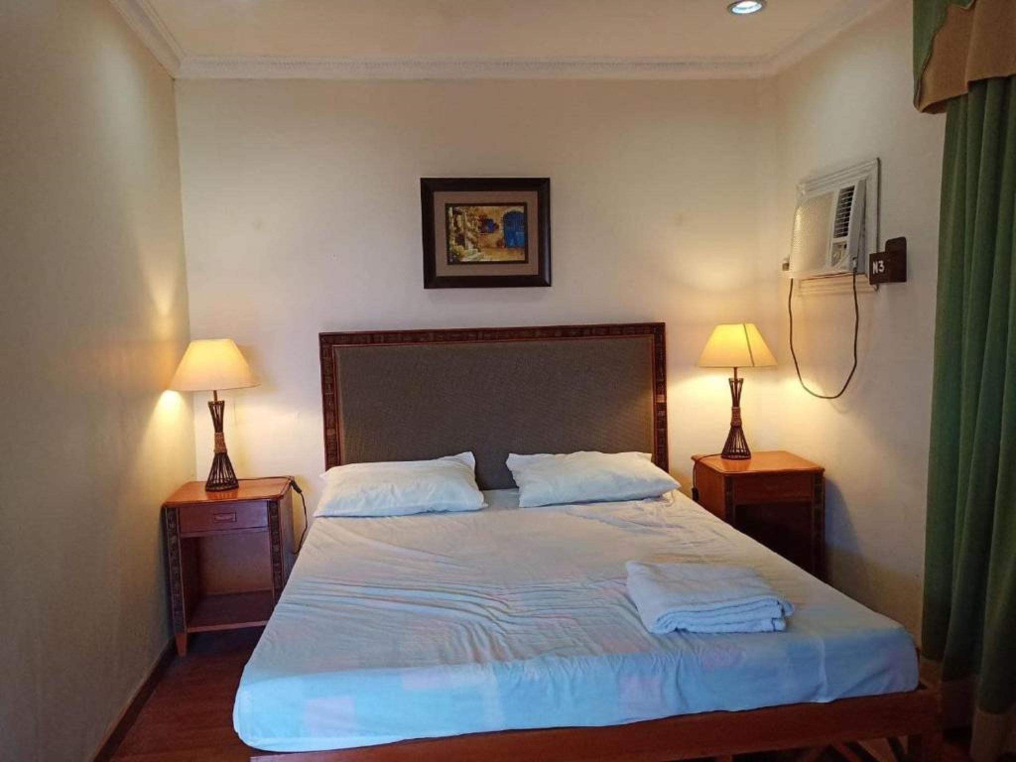 Quarto de hotel com uma cama grande, duas mesas de cabeceira com luminárias, um quadro na parede e uma janela com cortinas.