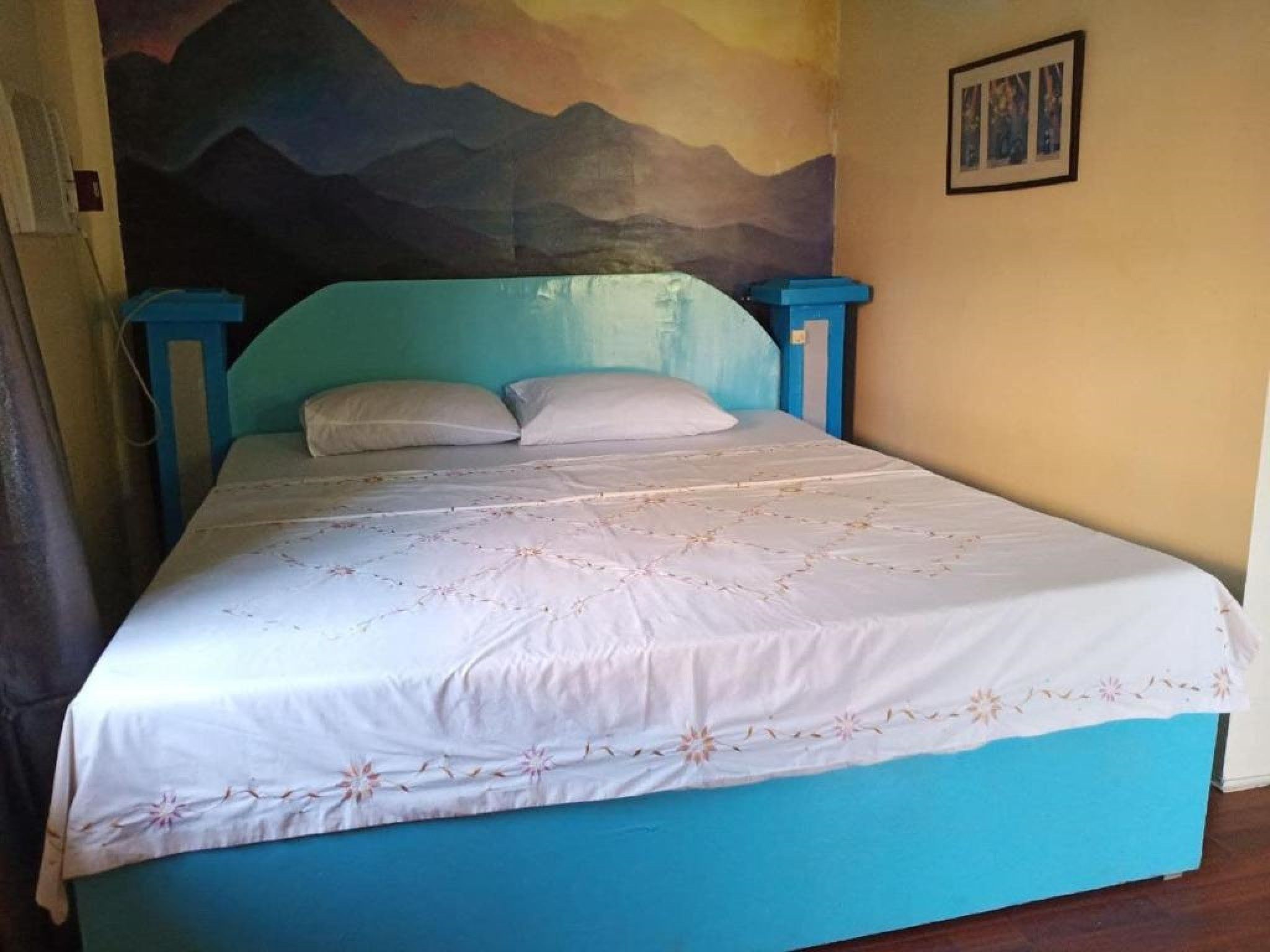 Uma cama com cabeceira azul e roupa de cama branca, um mural na parede e um quadro emoldurado.