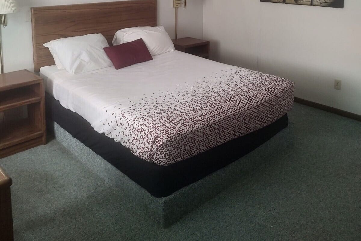 Quarto com cama de cabeceira de madeira, roupa de cama branca, manta estampada, relógio na parede e dois objetos decorativos em forma de animais.