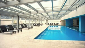 Piscina interna com teto de vidro, espreguiçadeiras e piso ladrilhado.