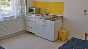 Cozinha compacta com micro-ondas, pia e armários. Há uma pequena lixeira amarela e uma mesa estampada com bolinhas com uma cadeira.