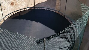 Um trampolim circular cercado por uma rede de proteção, com um muro de pedra e uma área de jardim ao fundo.