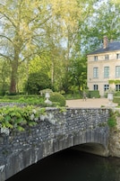 Uma ponte de pedra sobre a água, rodeada por vegetação exuberante e um edifício grande com um jardim.