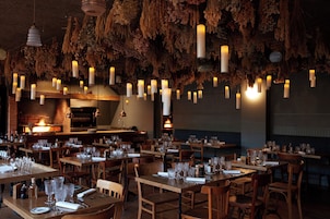 Restaurante com mesas e cadeiras de madeira, velas penduradas e parede de tijolos com lareira.