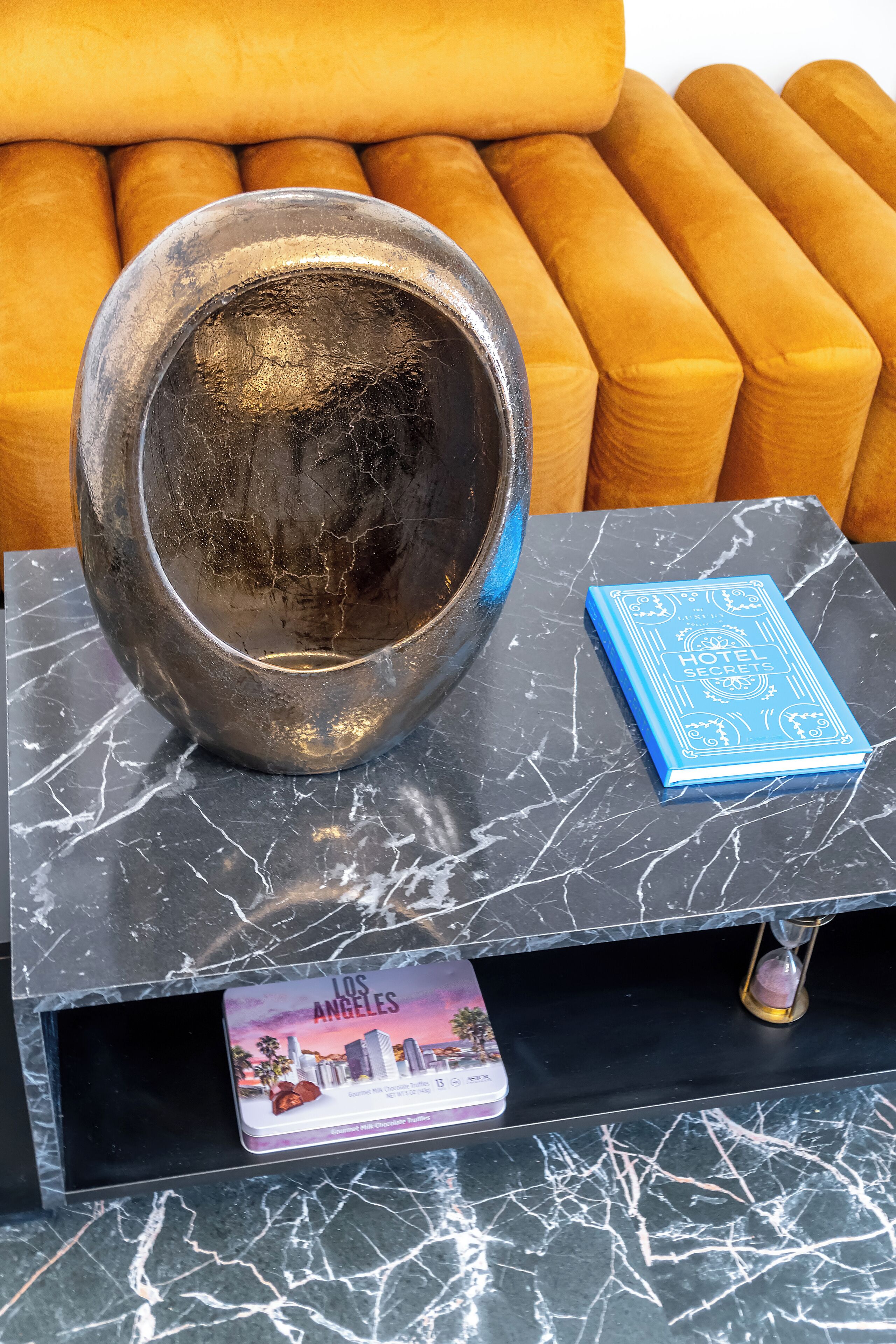Uma tigela metálica sobre uma mesa de mármore, um livro azul intitulado 'Hotel Secrets' e um cartão com tema de Los Angeles.