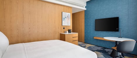 Quarto de hotel com uma cama grande, mesa de cabeceira de madeira, escrivaninha com cadeira, televisão de tela plana e uma parede azul decorativa.
