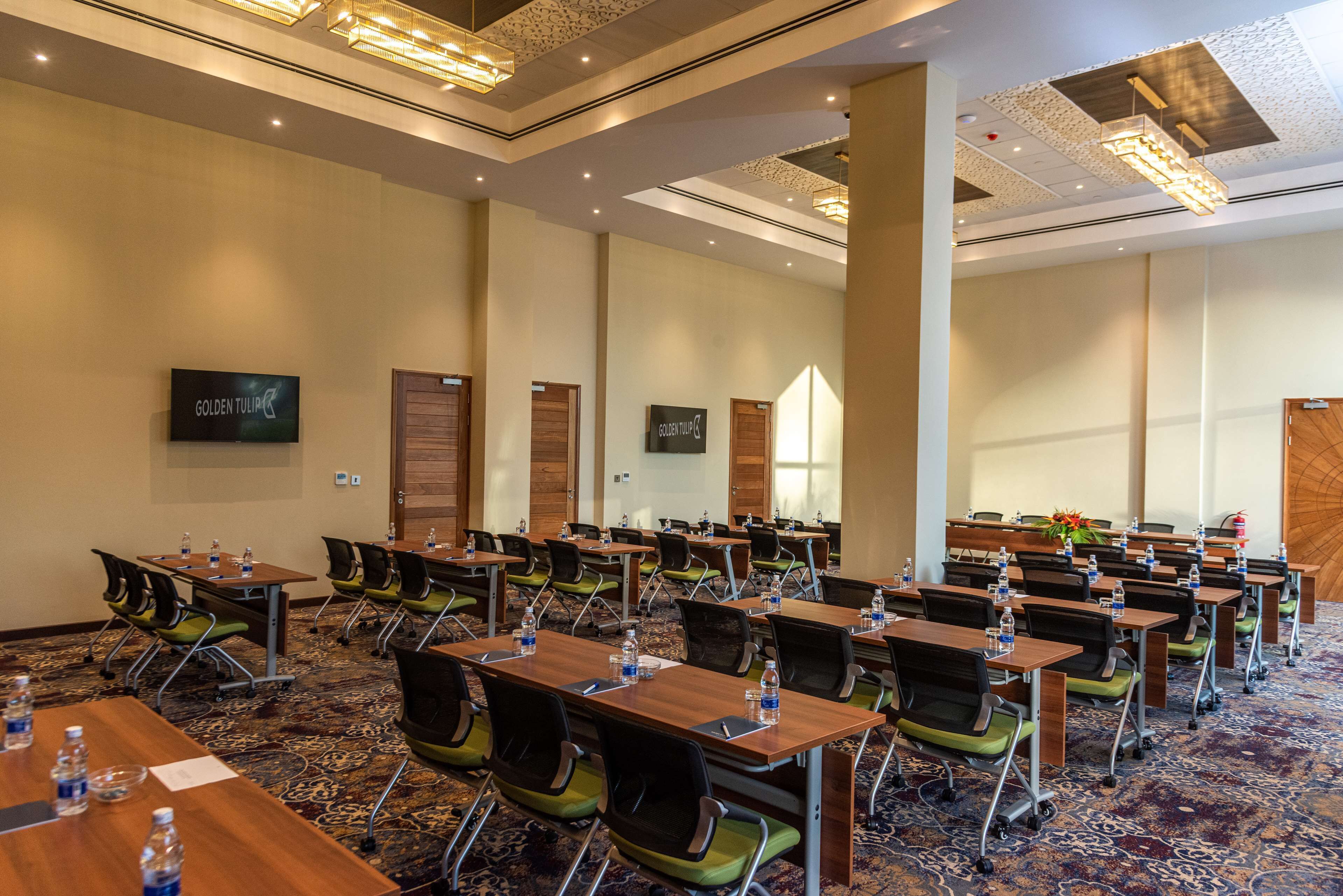 Uma sala de conferências com fileiras de mesas e cadeiras, equipada com garrafas de água e uma televisão na parede.