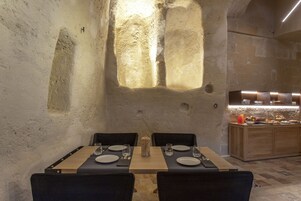 Uma área de refeições com uma parede de pedra rústica, uma mesa de madeira posta para dois e uma cozinha ao fundo.
