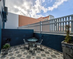 Um terraço no telhado com mesa e cadeiras, um vaso de planta e vista para um edifício ao fundo.