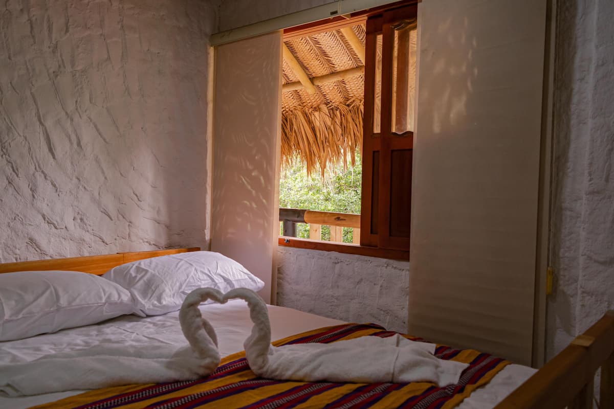 Quarto com cama, janela de madeira e telhado de palha visível pela janela.