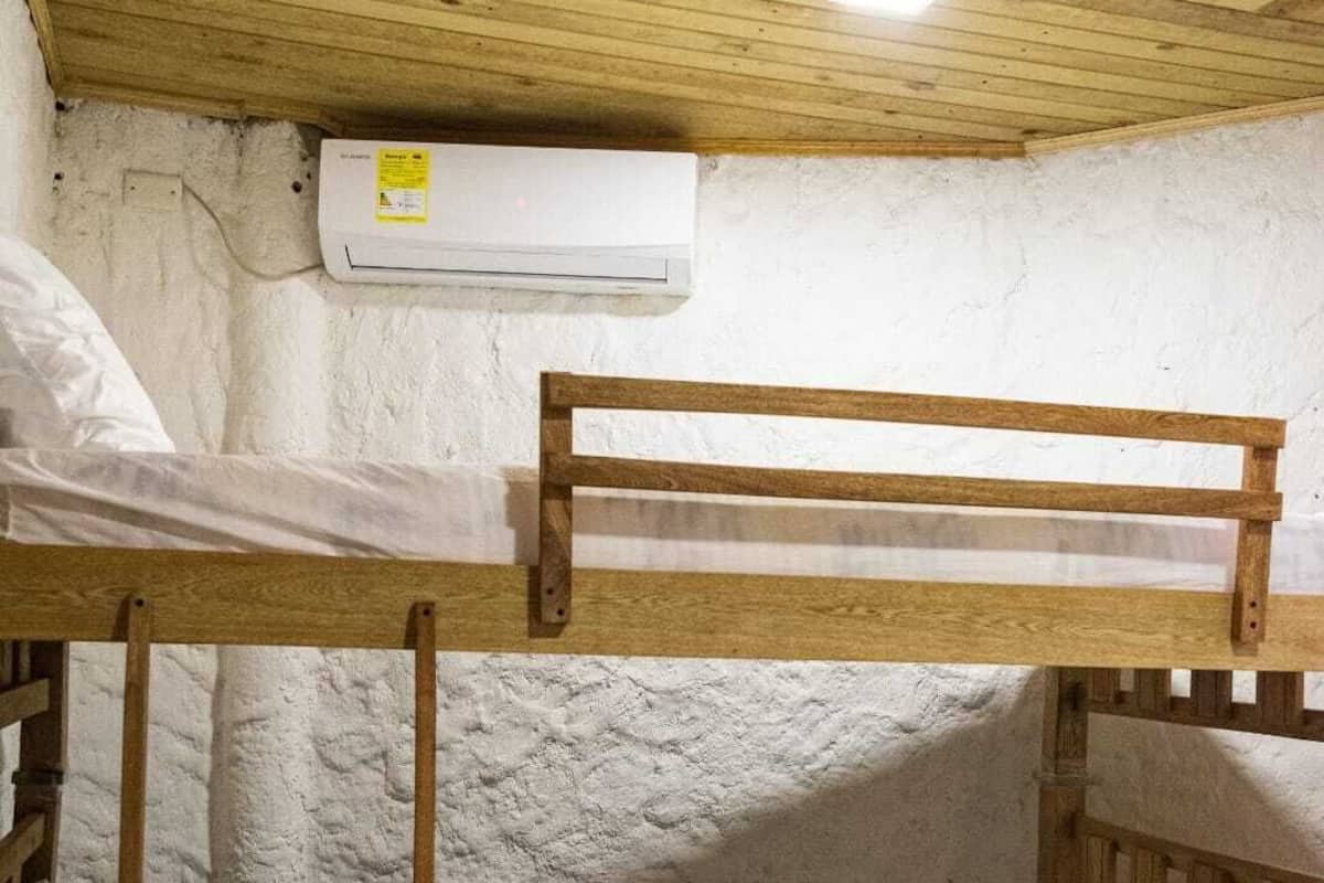Quarto com beliche, escada de madeira, paredes brancas e aparelho de ar condicionado.