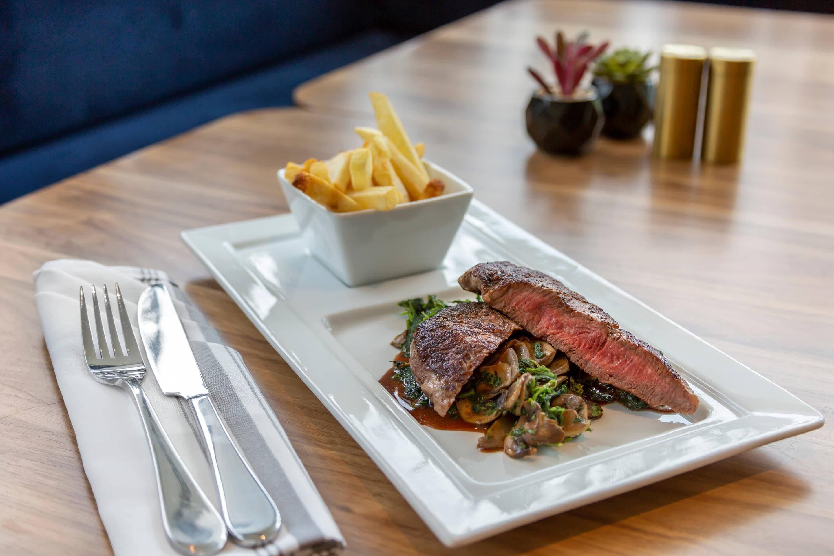 Um prato de bife com cogumelos e batatas fritas sobre um prato branco retangular, acompanhado de talheres e guardanapo em uma mesa de madeira.