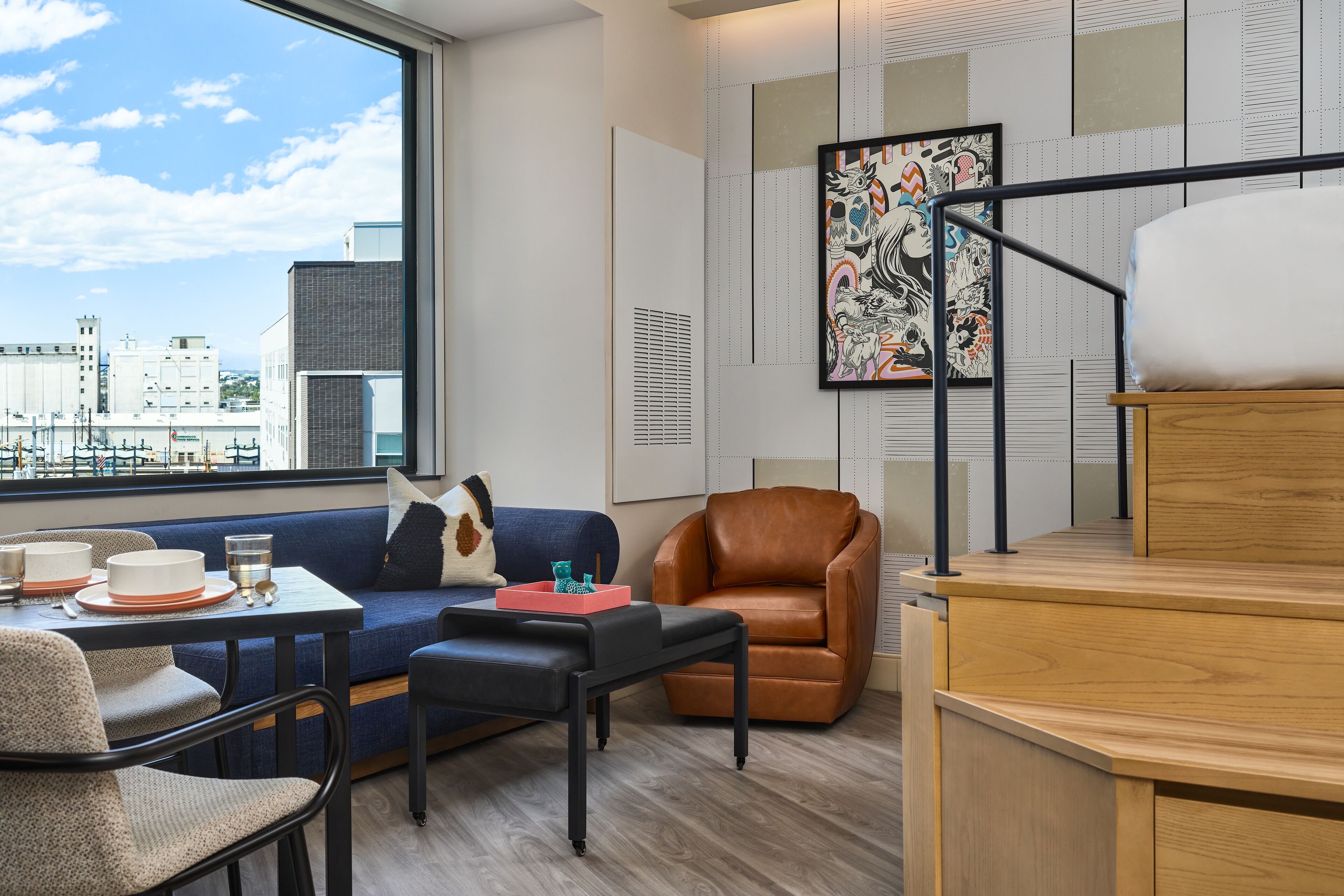 Uma sala de estar moderna com um sofá, uma poltrona de couro marrom, uma mesa de jantar com pratos e copos, e uma janela grande com vista para a cidade.
