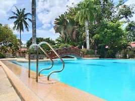 Piscina com uma ponte de pedra, rodeada por palmeiras e um céu claro.