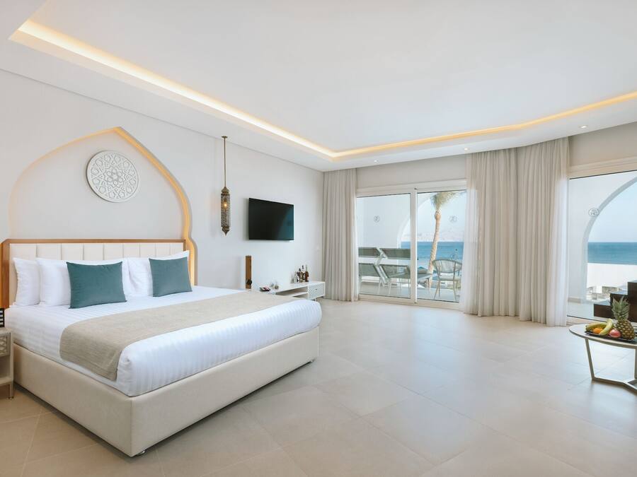 Quarto de hotel moderno com uma cama grande, TV, varanda com vista para o mar e uma pequena mesa com frutas.