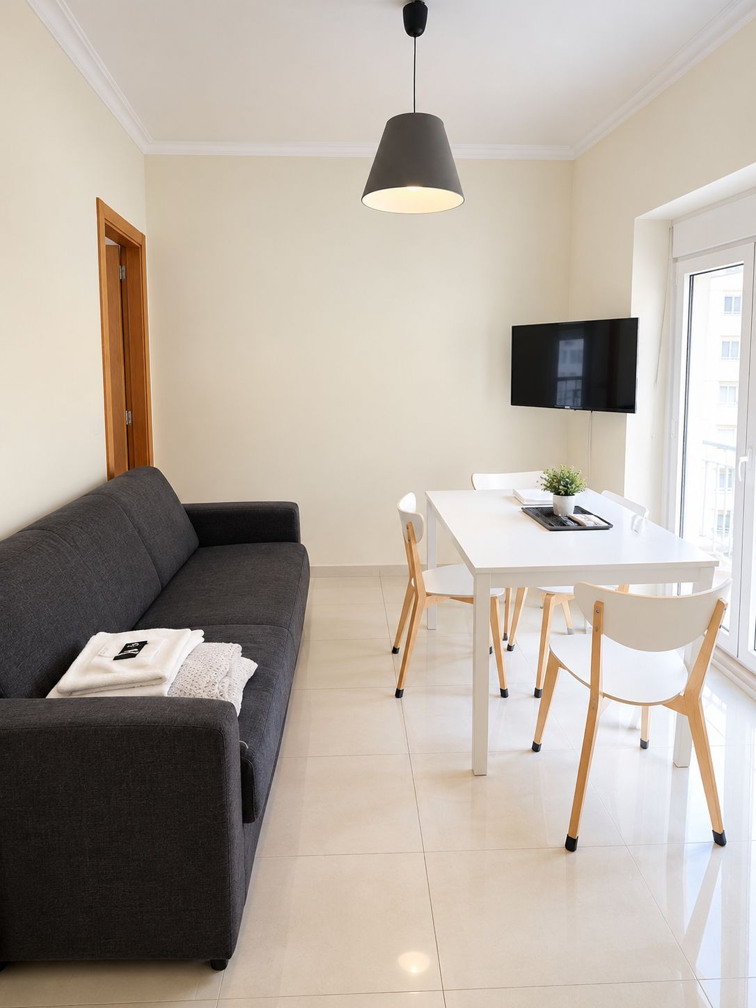 Uma sala de estar moderna com um sofá cinzento, uma mesa de jantar branca com cadeiras de madeira, uma luminária pendente preta e uma TV de tela plana fixada na parede.