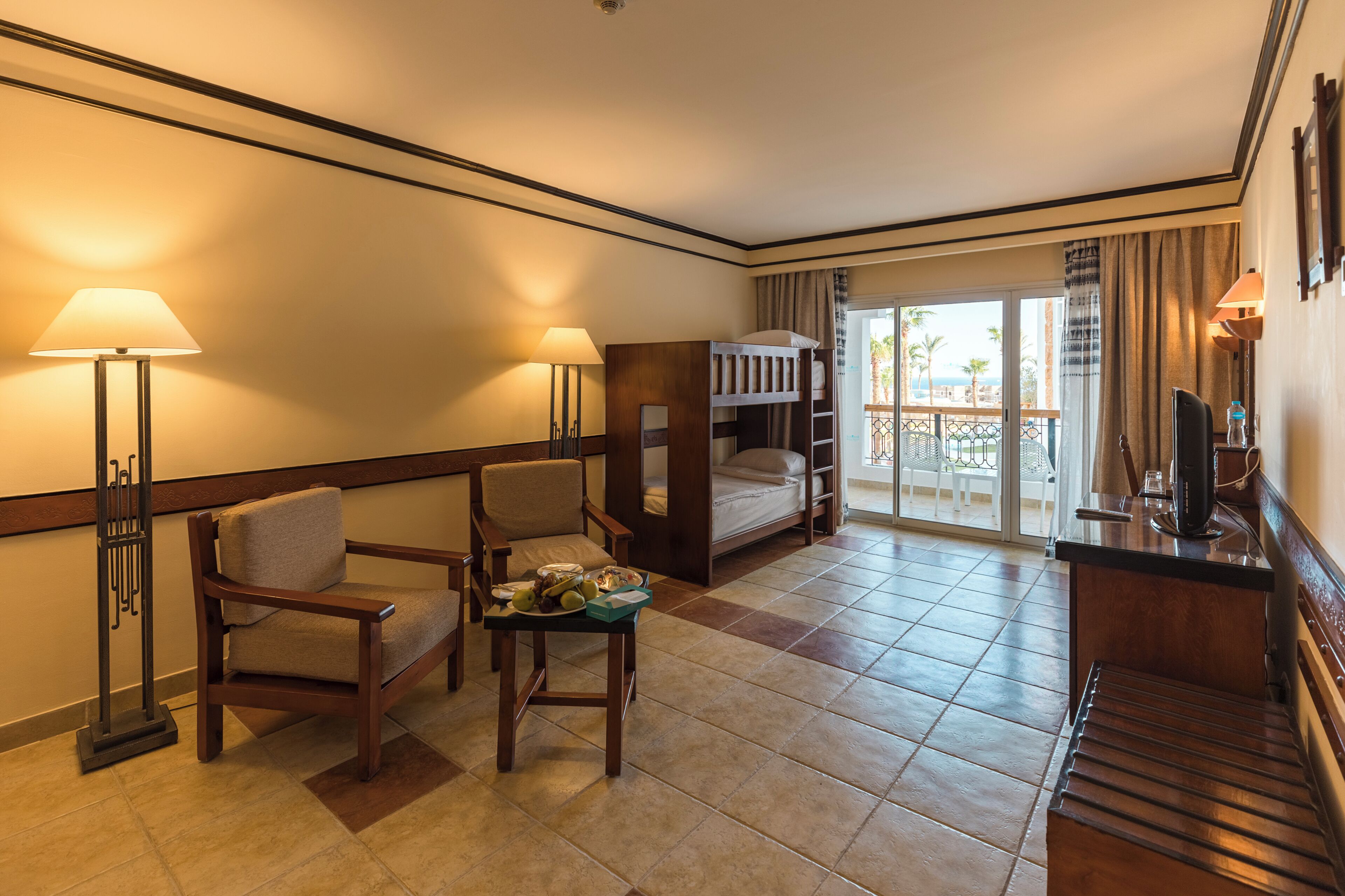 Quarto de hotel com cama, escrivaninha, cadeira, mesa com frutas e varanda com vista.