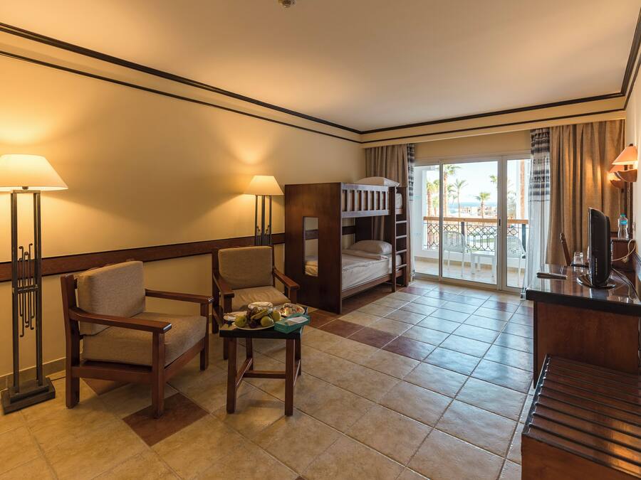 Quarto de hotel com cama, escrivaninha, cadeira, mesa com frutas e varanda com vista.
