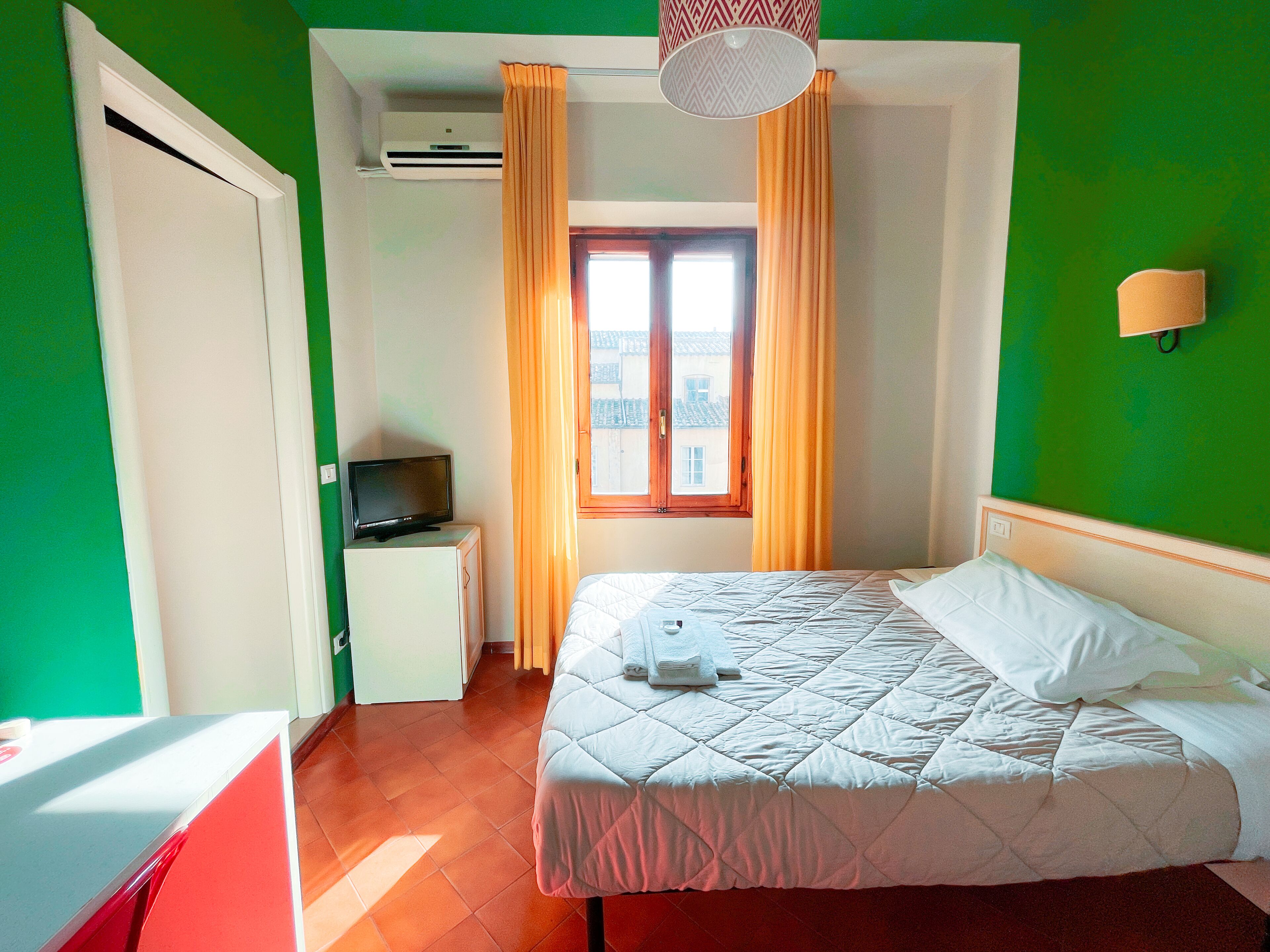 Quarto de hotel com uma cama, televisão sobre um móvel pequeno, janela com cortinas e uma luminária na parede.