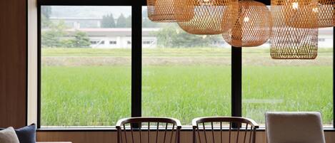 Uma área de refeições com uma grande janela, luminárias pendentes de vime e vista para um campo verde.