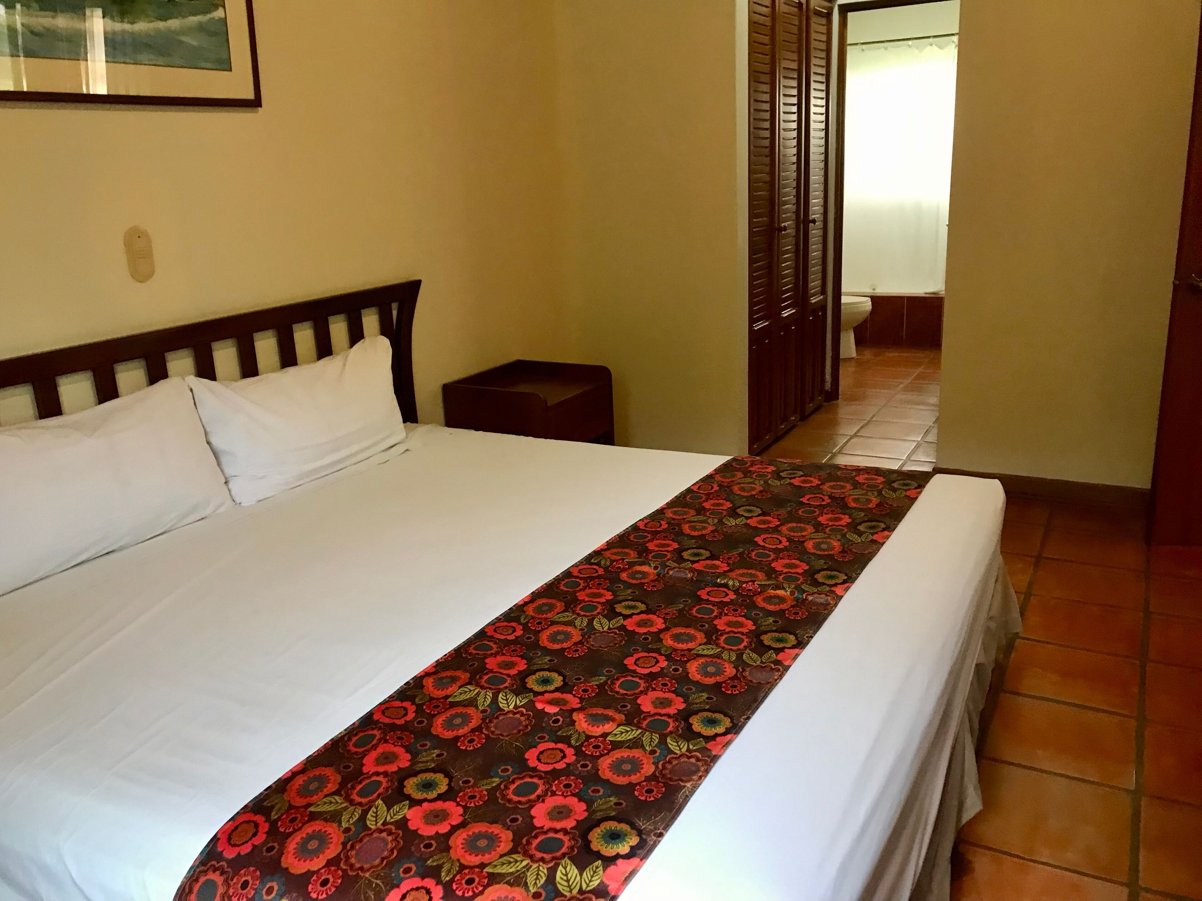 Quarto de hotel com cama, mesa de cabeceira, cabeceira de madeira, porta e chão ladrilhado.