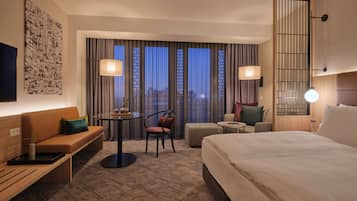 Quarto de hotel com uma cama grande, um sofá, uma mesinha e uma cadeira. Cortinas e vista para a cidade através das janelas.