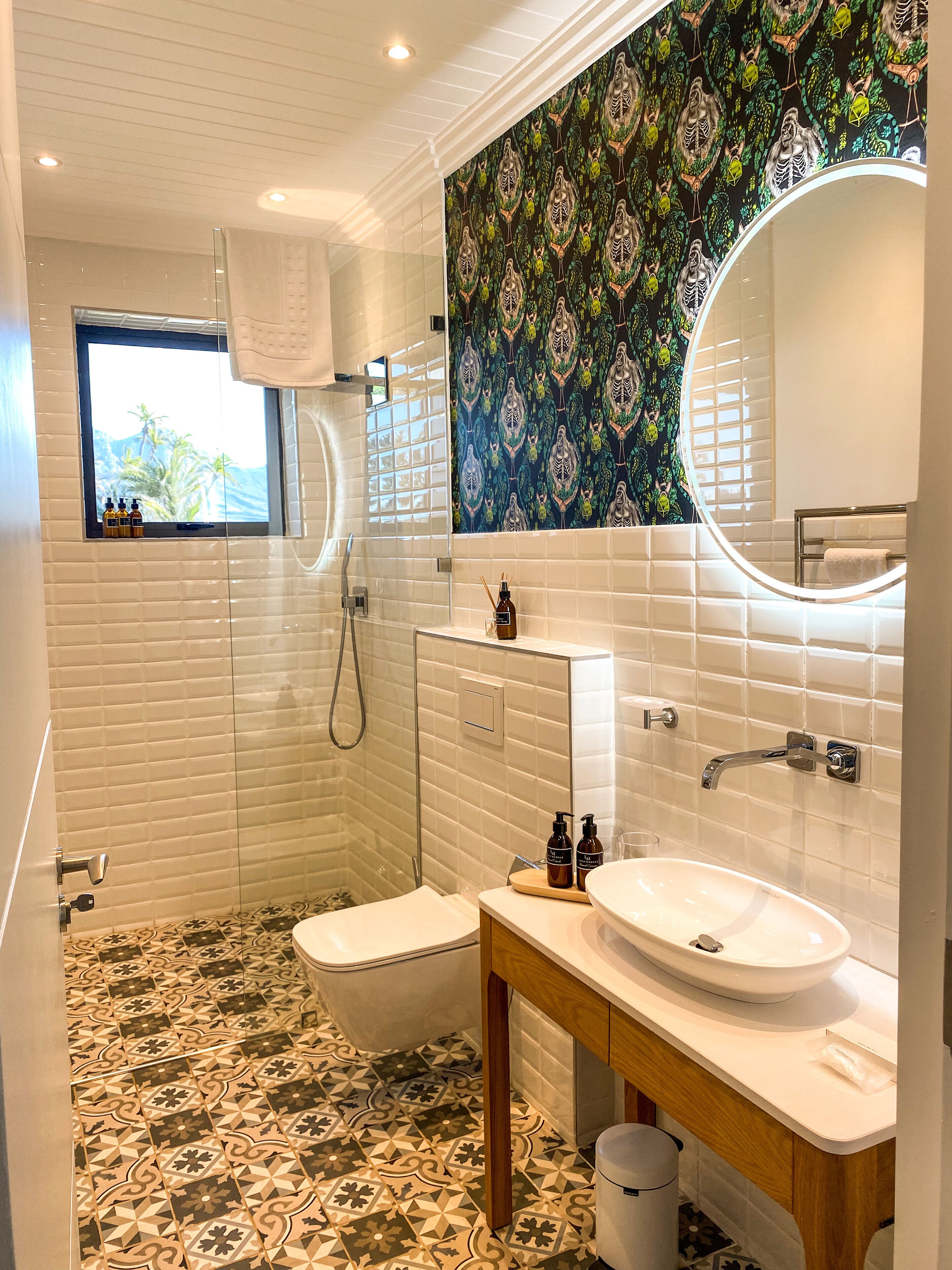 Um banheiro moderno com vaso sanitário, pia e chuveiro. Apresenta azulejos estampados, uma janela com vista e um espelho decorativo.