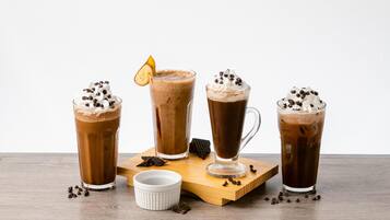 Quatro copos de café gelado com chantilly e raspas de chocolate, dispostos numa tábua de madeira com uma pequena tigela branca e chocolate picado espalhado.