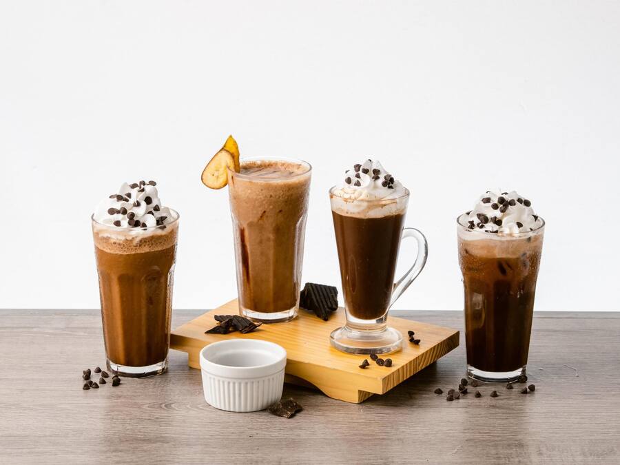 Quatro copos de café gelado com chantilly e raspas de chocolate, dispostos numa tábua de madeira com uma pequena tigela branca e chocolate picado espalhado.