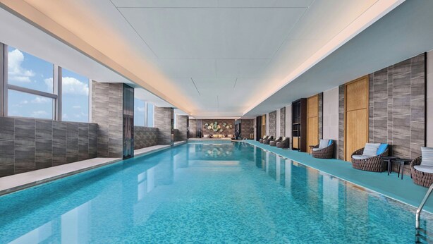 Uma piscina interior extensa com água azul cristalina, rodeada por paredes com texturas mistas de madeira e pedra, e áreas de estar com mobiliário de vime.