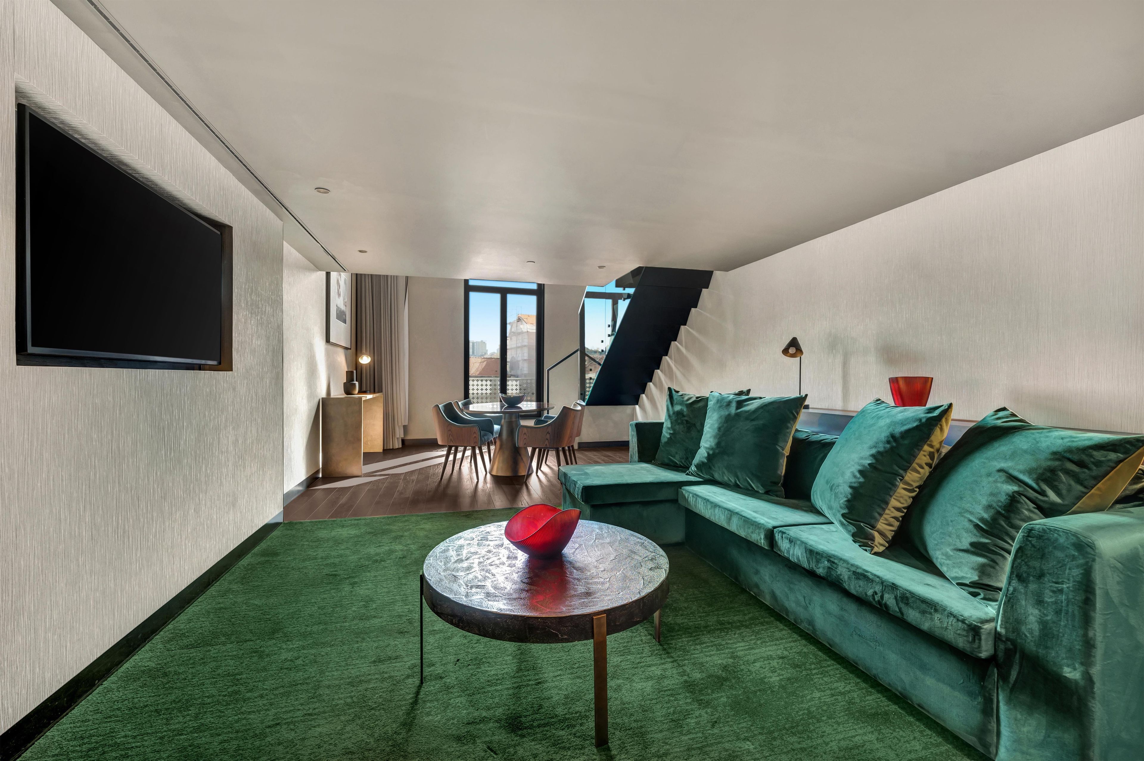 Uma sala de estar moderna com carpete verde, uma mesa de centro redonda, um sofá verde com almofadas, uma TV de tela plana fixada na parede e uma escada que leva a um andar superior.