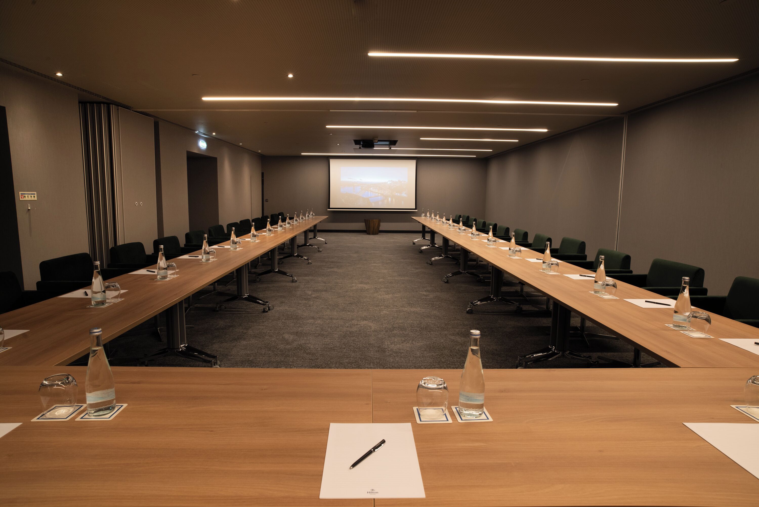 Sala de conferências com mesas compridas, cadeiras, garrafas de água e uma tela de projetor.