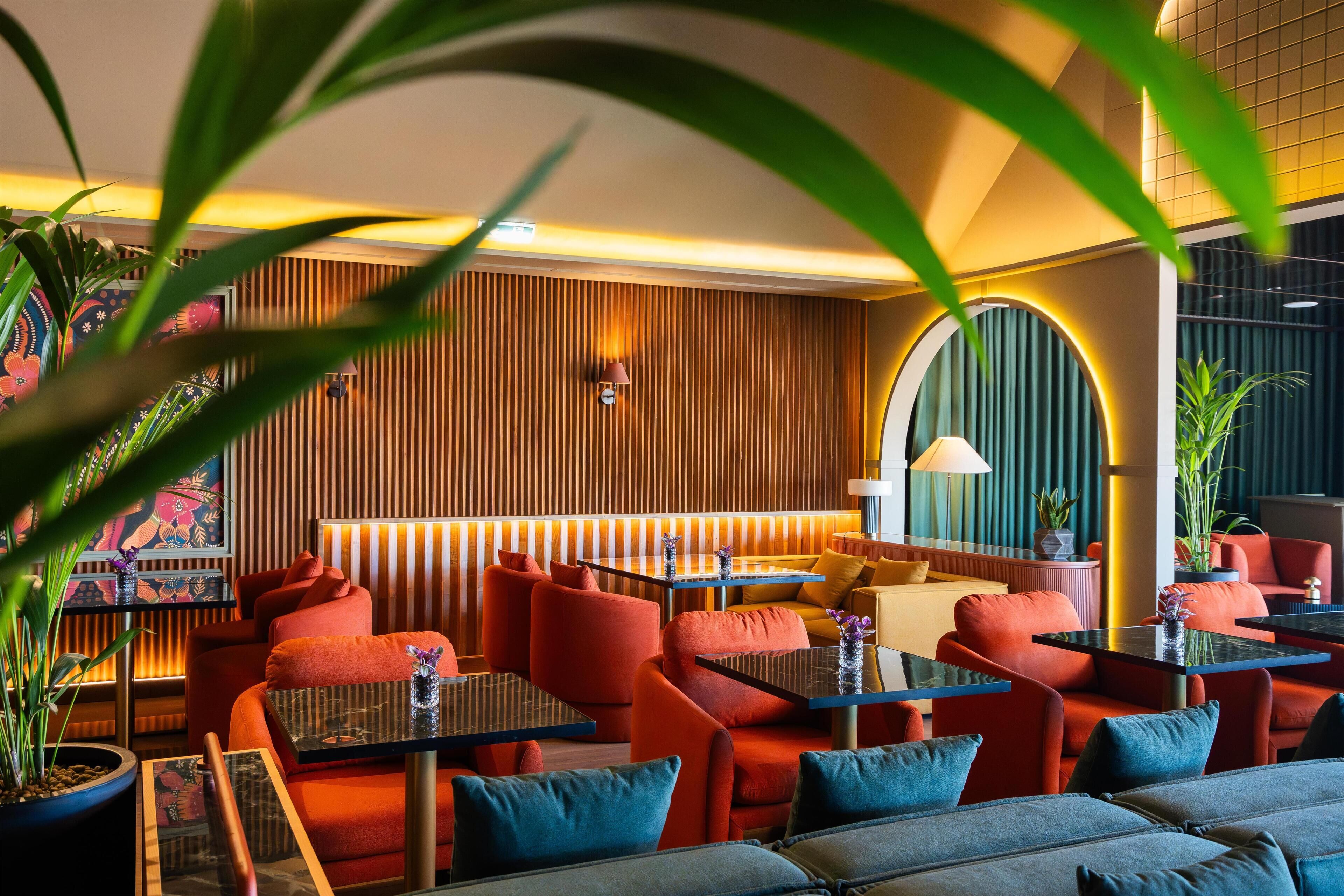 Uma área de estar moderna, com assentos em vermelho e azul, uma parede de destaque em madeira e uma mesa redonda com abajur.