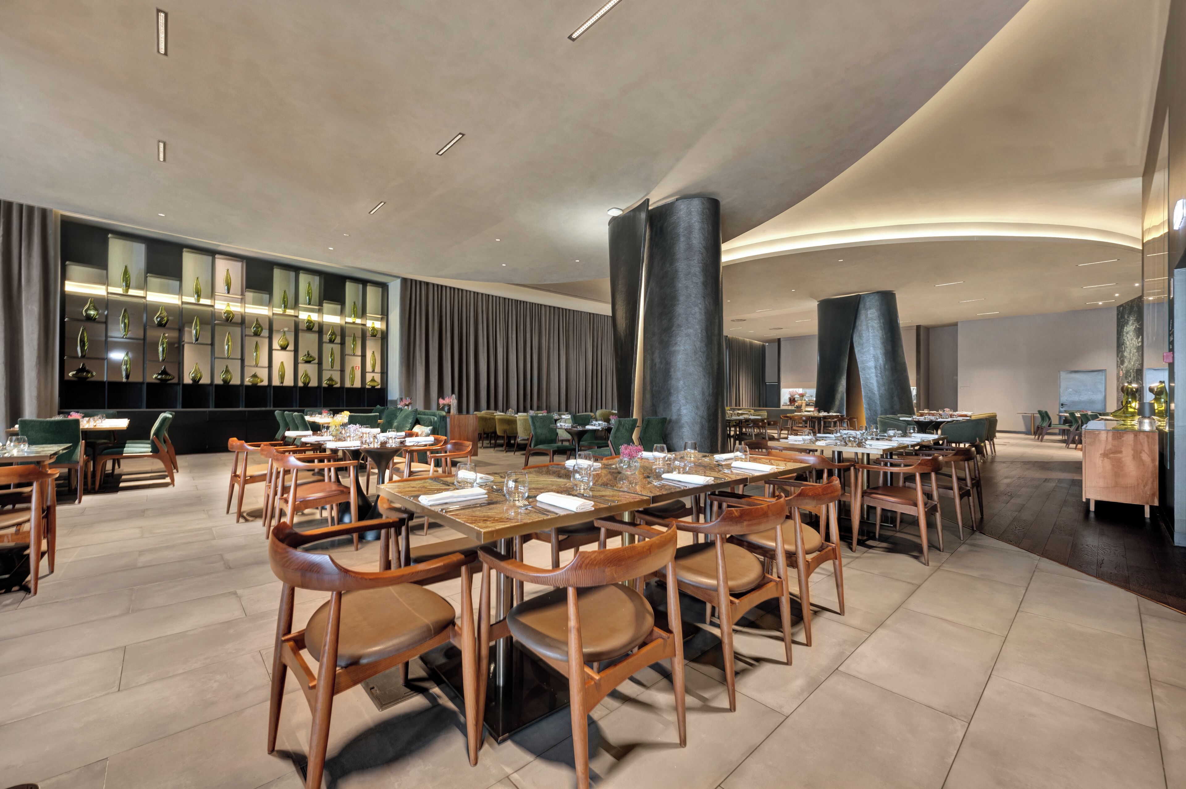 Uma ampla área de refeições com várias mesas e cadeiras, um bar com copos e taças, e um design moderno no teto.