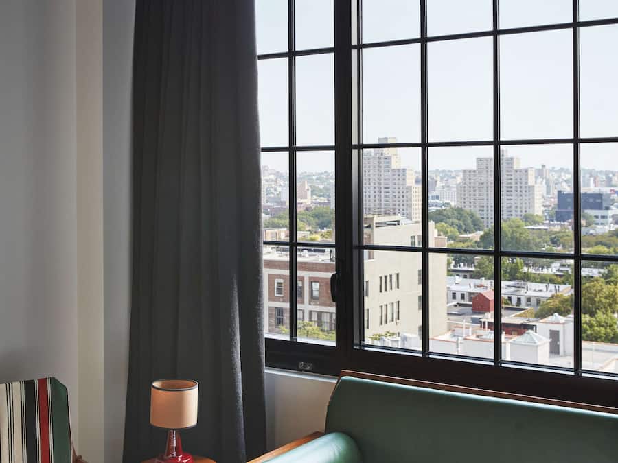 Quarto de canto com sofá verde, cadeira listrada e um abajur perto de uma janela com vista para a cidade.