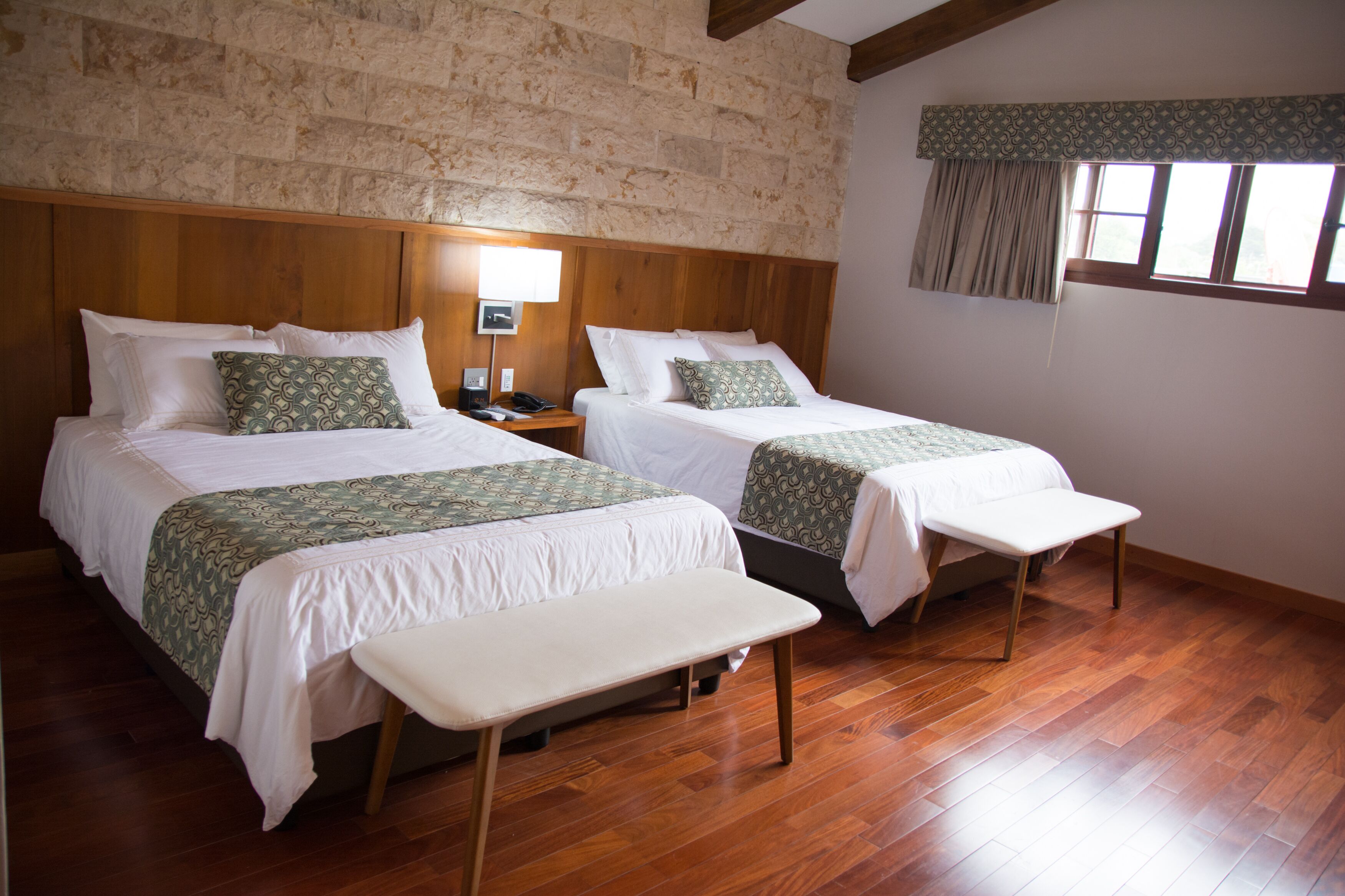 Quarto com duas camas, piso de madeira, parede de tijolos e janela com cortinas.