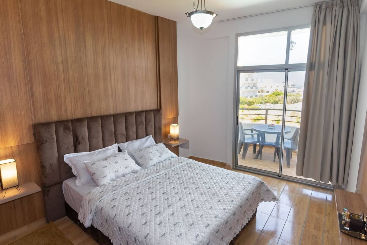 Quarto com cama grande, cabeceira de madeira, abajures e vista para um terraço ao ar livre com mesa e cadeiras.