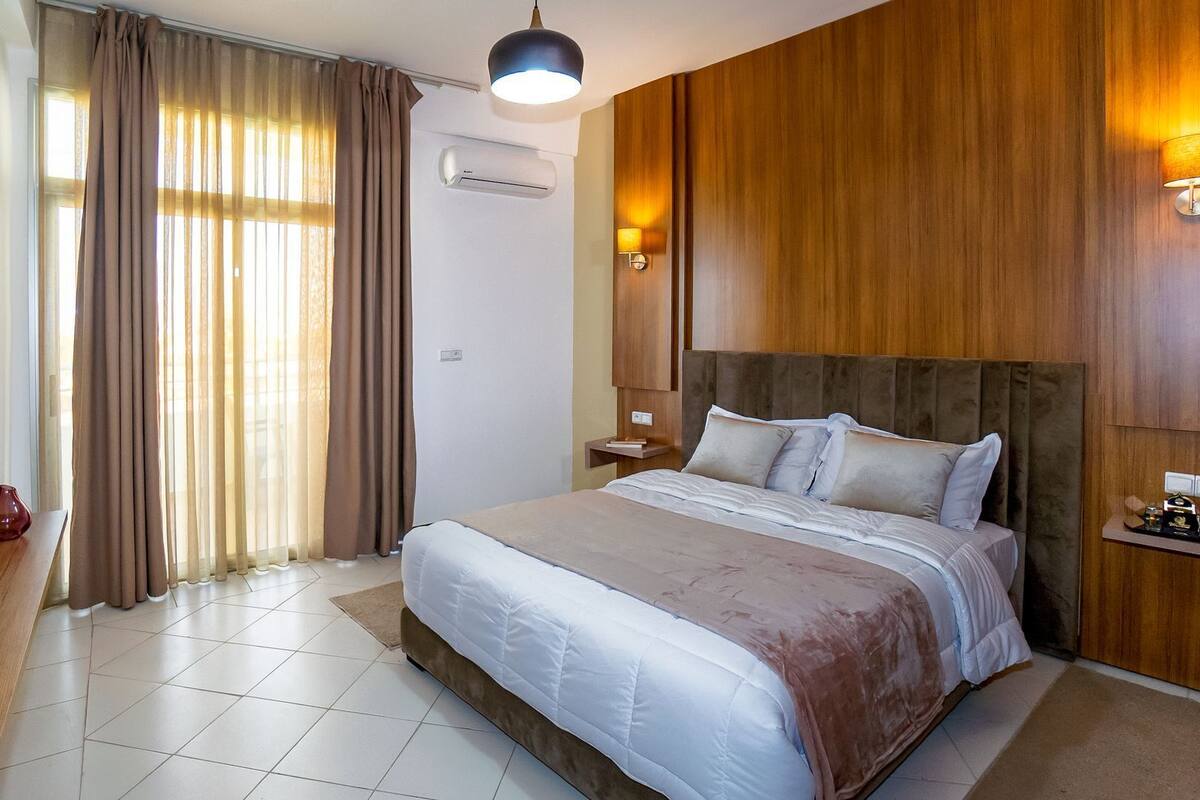 Quarto de hotel com uma cama grande, painéis de madeira, luminária de cabeceira e uma janela com cortinas.