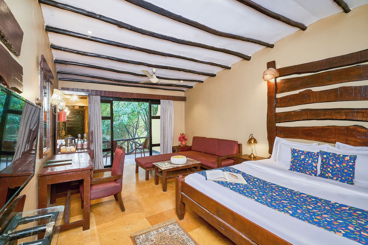 Quarto com cama, sofá, mesa e cadeira. Há uma janela com cortinas, um ventilador de teto e vista para árvores.