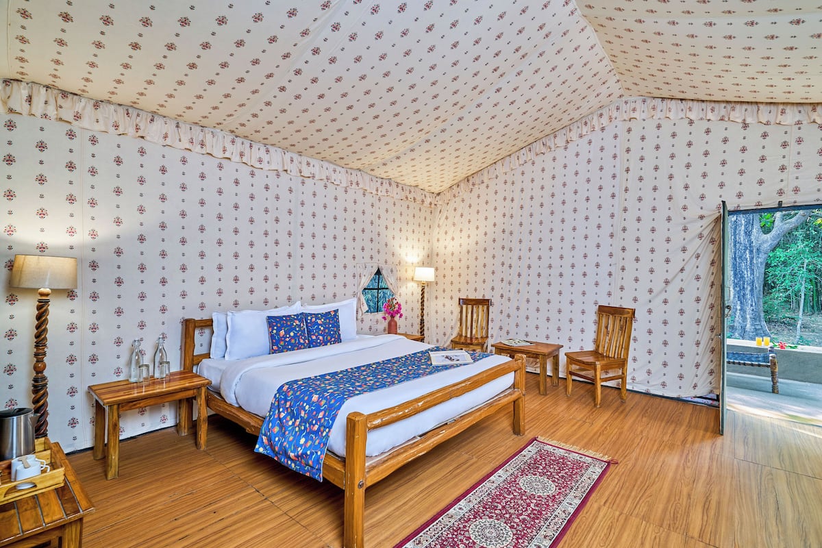Quarto com cama de madeira, mesa de cabeceira, cadeira, abajur e janela com vista para árvores.