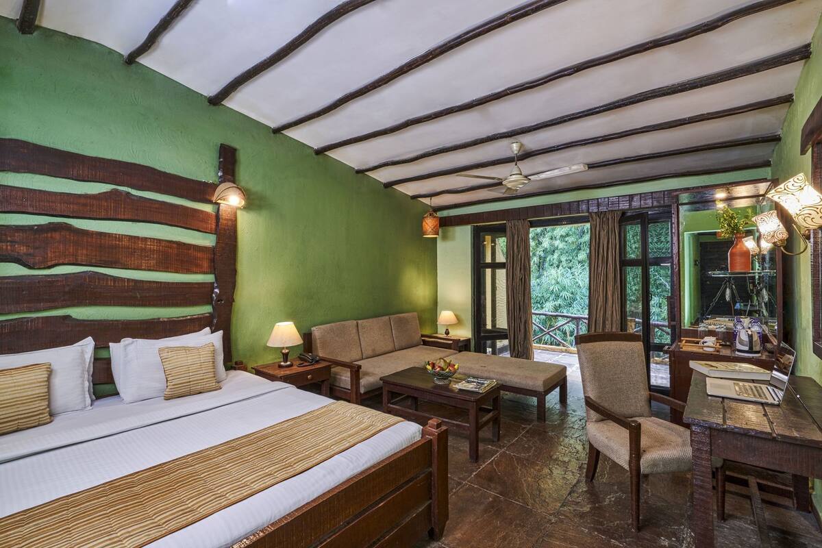 Quarto com cama, sofá e poltrona, uma mesinha com abajur e vista para o verde lá fora.