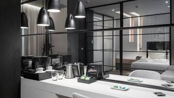 Quarto de hotel moderno com um espelho grande, uma escrivaninha branca com cadeira e uma área de kitchenette.