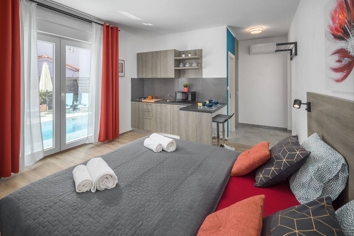 Quarto de hotel moderno com cama, pequeno espaço de cozinha e porta de vidro deslizante para o exterior.