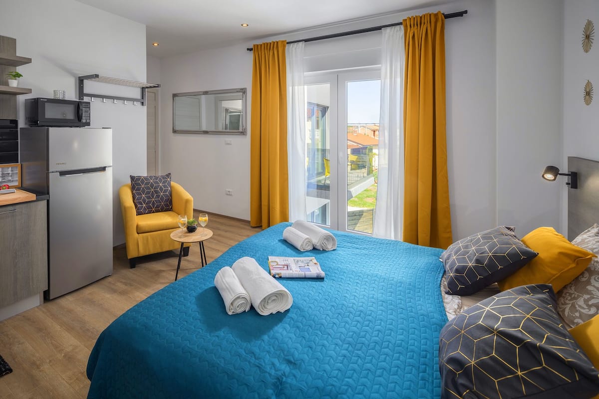 Quarto de hotel com uma cama, uma cadeira, uma mesinha, micro-ondas e geladeira.